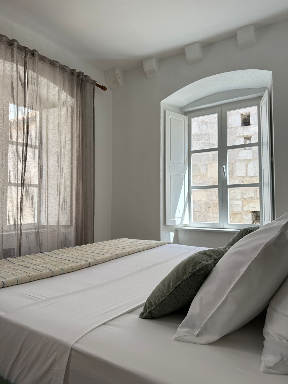 Quarto com cama, janela com paredes de pedra e cortinas leves.