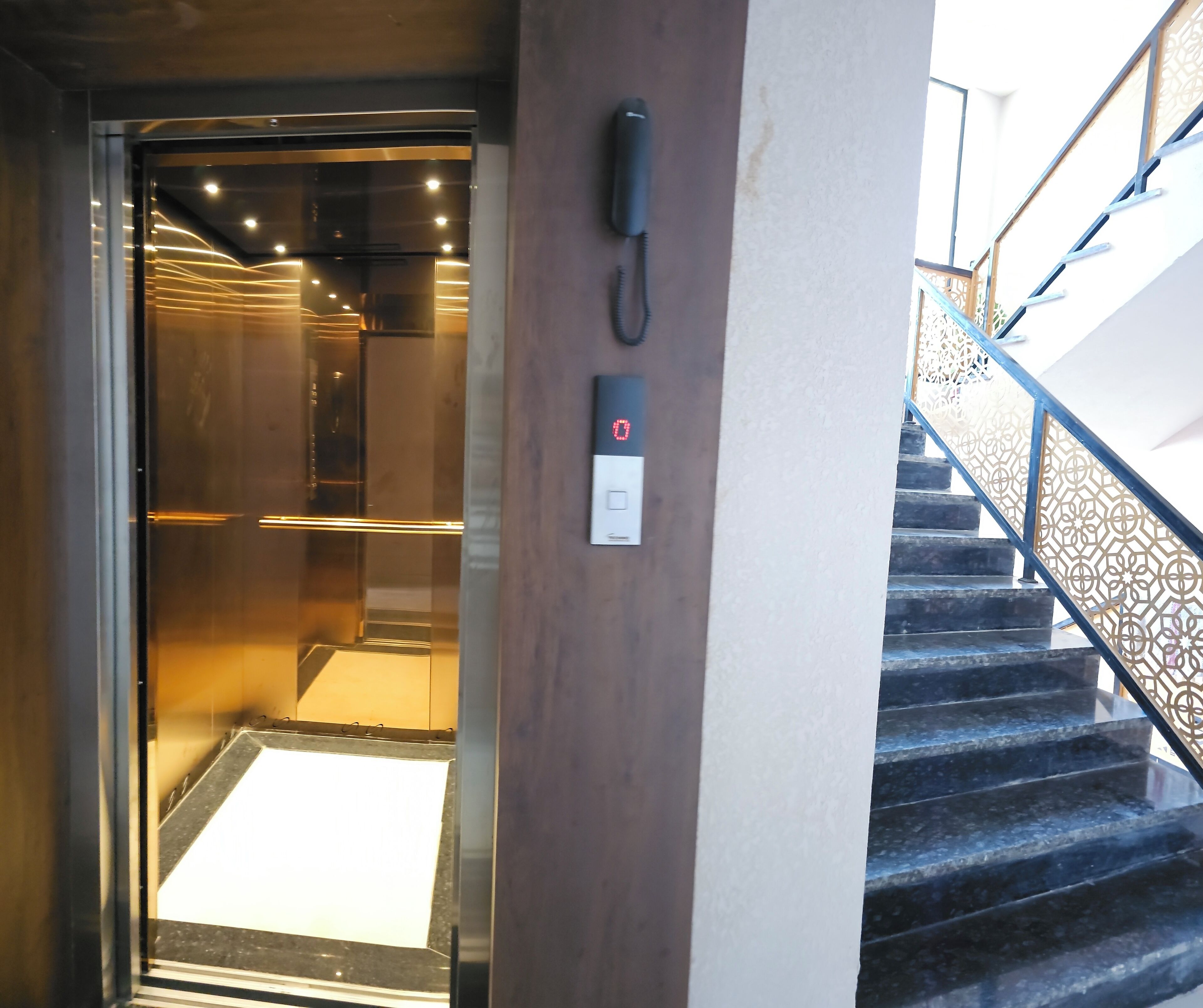 Interior de elevador com painel digital, interfone fixado na parede e escada com corrimão decorativo.