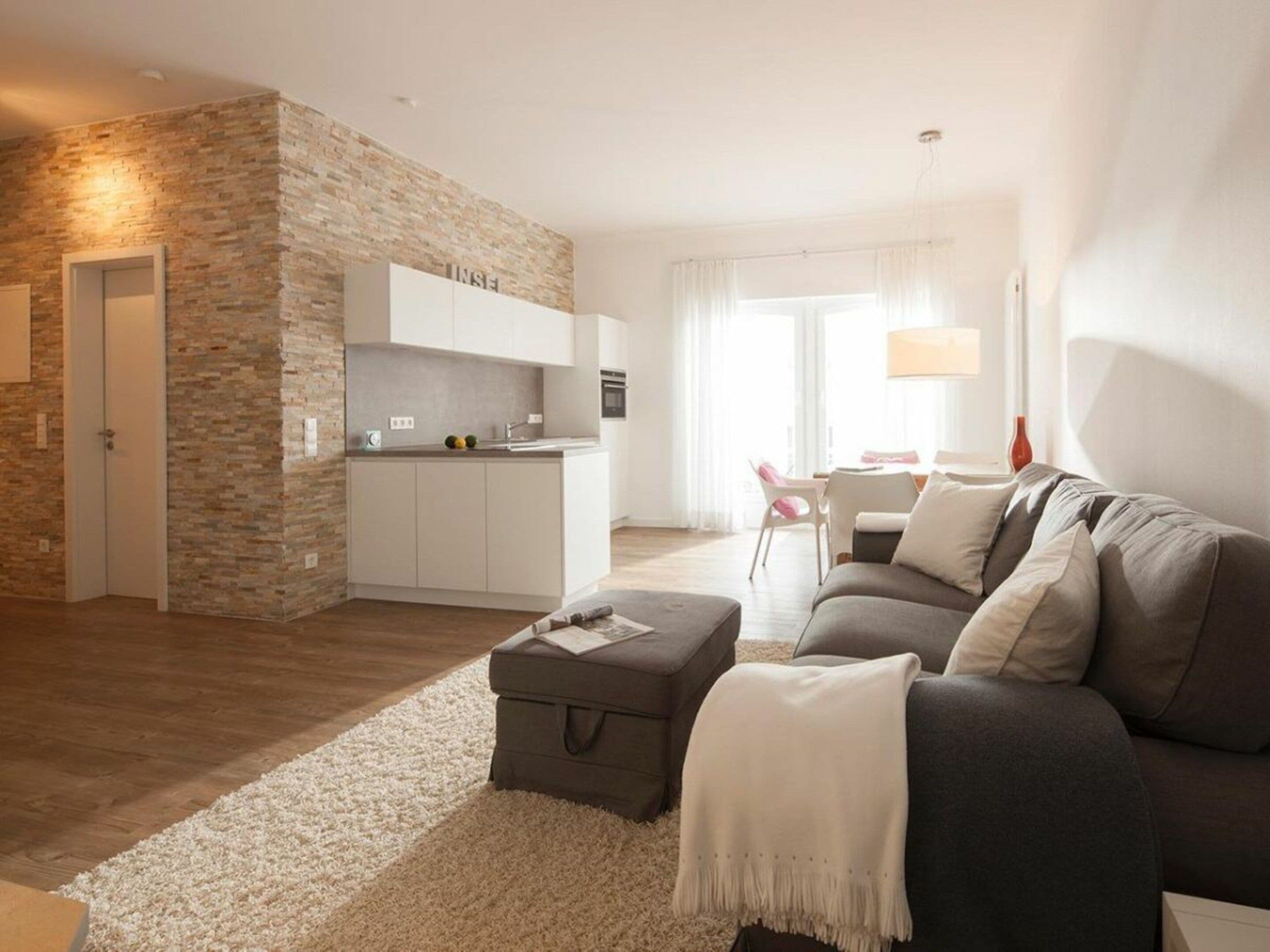 Apartamento moderno com parede de destaque em pedra, cozinha compacta, área de refeições e sala de estar com sofá cinza e manta branca.