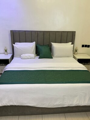 Cama bem arrumada com cabeceira cinza, travesseiros brancos e verdes, e colcha verde.