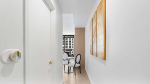 Um corredor com uma porta branca, uma área de refeições com mesa e cadeiras, e vista para um edifício através de uma janela.