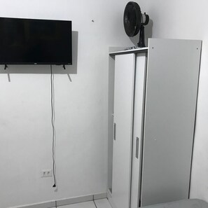 Quarto com TV de tela plana fixada na parede, guarda-roupa branco com um ventilador em cima e uma tomada visível na parede.