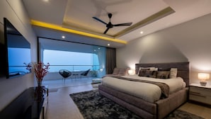 Quarto moderno com cama grande, ventilador de teto, televisão e varanda com vista para o mar.