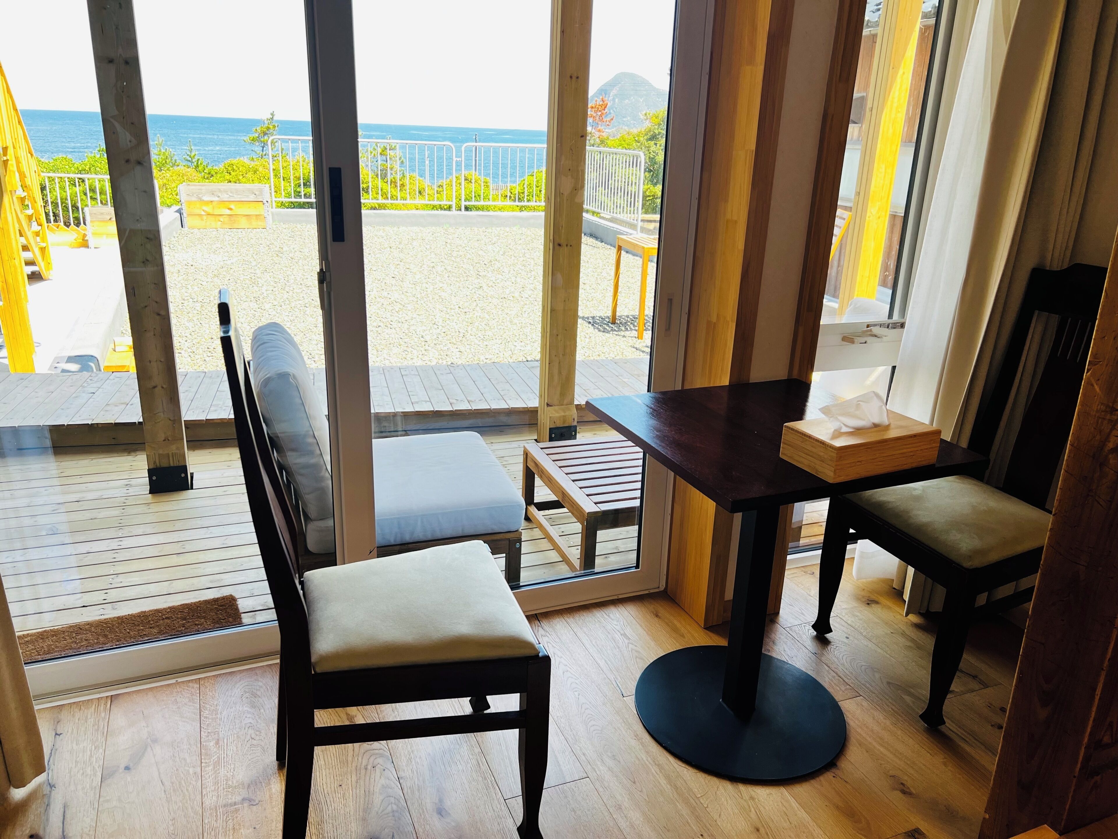 Quarto com piso de madeira, sofá, mesa e vista para o mar através de grandes janelas.