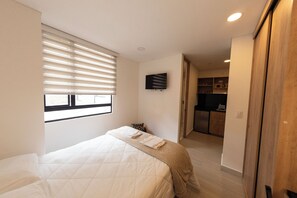 Quarto com cama, janela com persianas, televisão na parede e área de cozinha ao fundo.