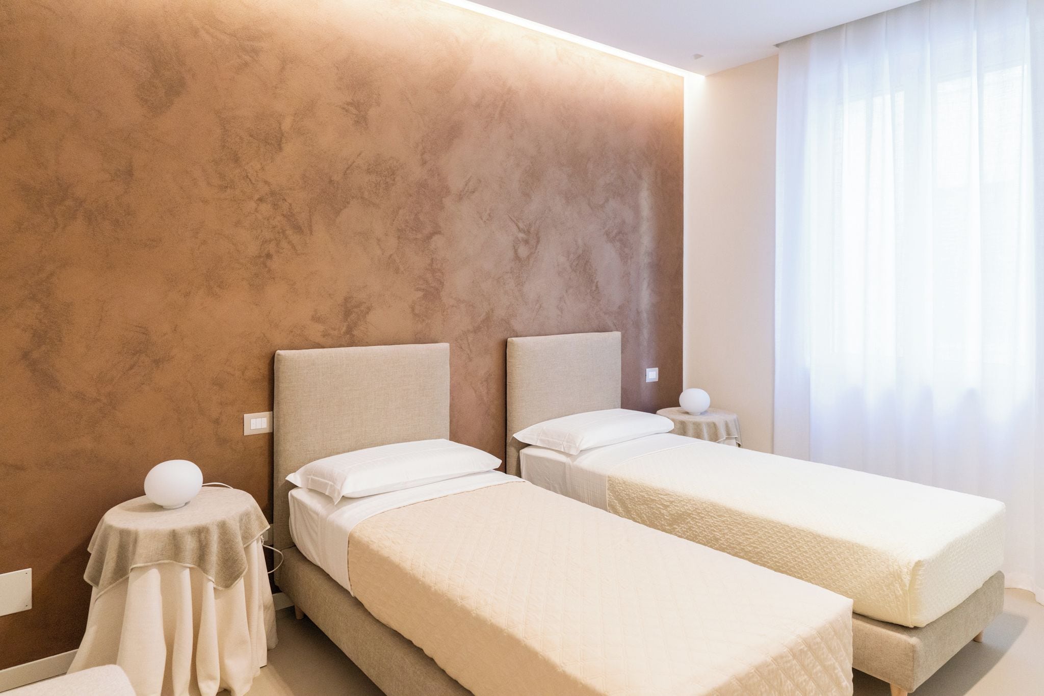 Quarto de hotel com duas camas, uma mesa redonda com abajur e uma janela com cortinas.