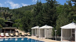 Piscina com espreguiçadeiras e toldos, cercada por árvores e uma colina.