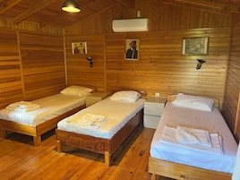Uma cabana com três camas de madeira, cada uma com travesseiro e toalha dobrada. Há luminárias fixadas na parede e um ventilador de teto.