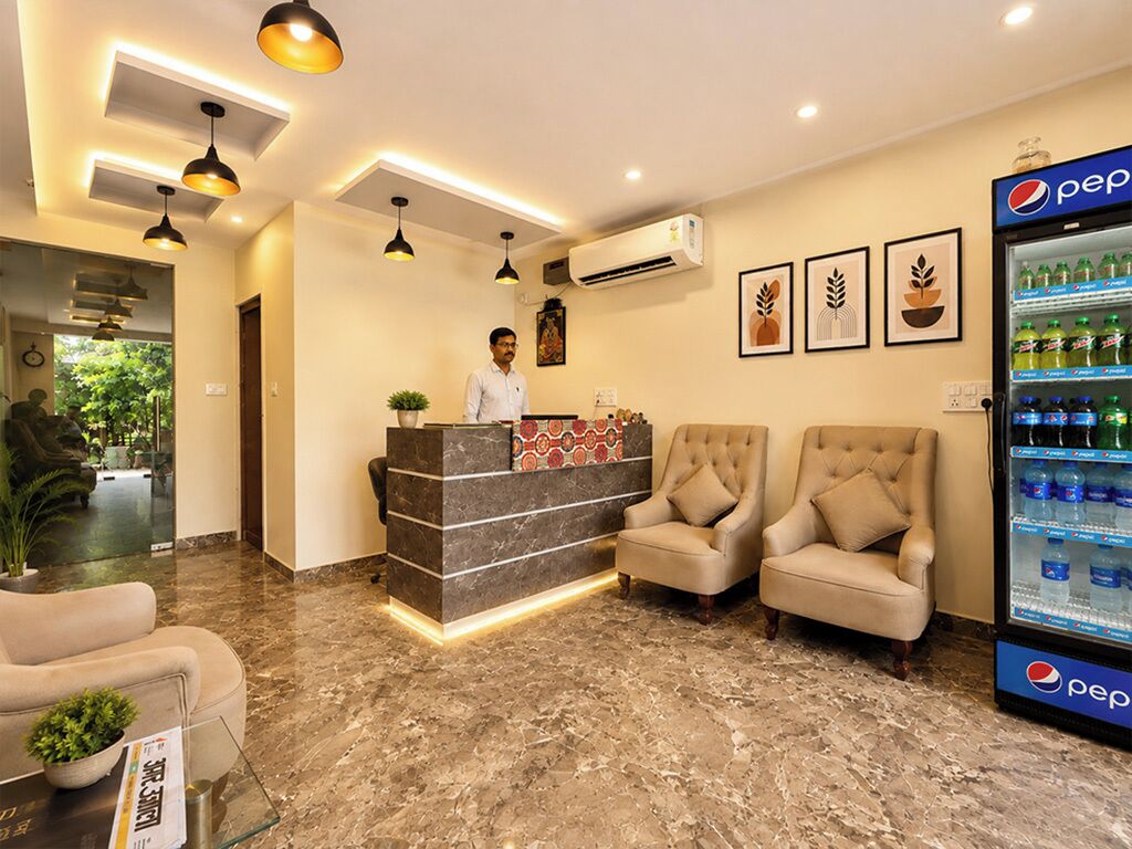 Uma área de recepção de hotel com piso de mármore, um balcão de atendimento, duas poltronas, um refrigerador da Pepsi e um homem parado atrás do balcão.