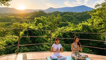 Duas pessoas praticando yoga em tapetes ao ar livre, com uma vista panorâmica de uma floresta e montanhas ao fundo.