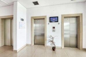 Área de elevadores limpa e moderna, com três elevadores, um visor digital e uma planta em vaso.