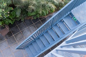 Escada de metal azul com um sinal de saída de emergência verde, rodeada por vegetação e um chão de azulejos.