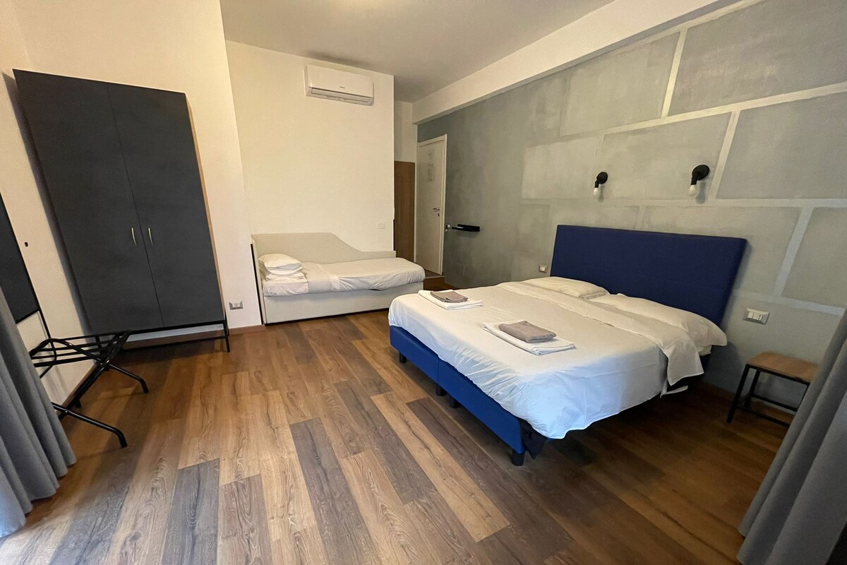 Quarto de hotel com cama, uma cadeira, guarda-roupa e porta.