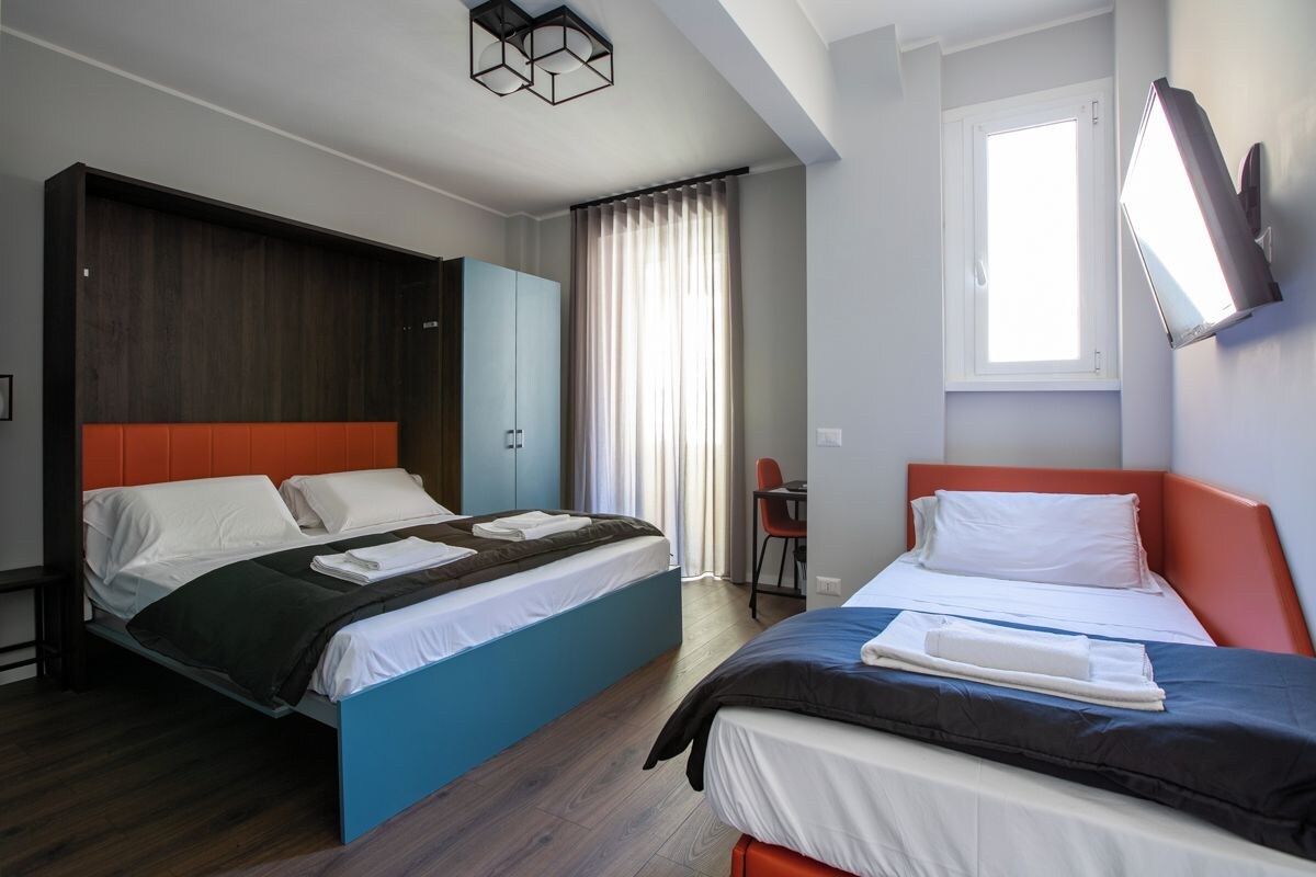 Quarto de hotel com duas camas, uma escrivaninha, uma cadeira e uma televisão na parede.