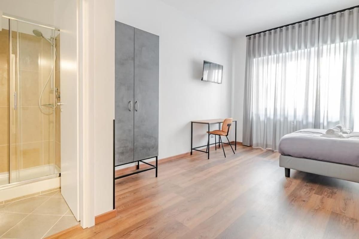 Quarto moderno com cama, escrivaninha, cadeira, armário e área de chuveiro.