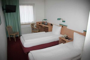 Quarto de hotel com duas camas, uma escrivaninha, uma cadeira, uma televisão e uma janela com cortinas.