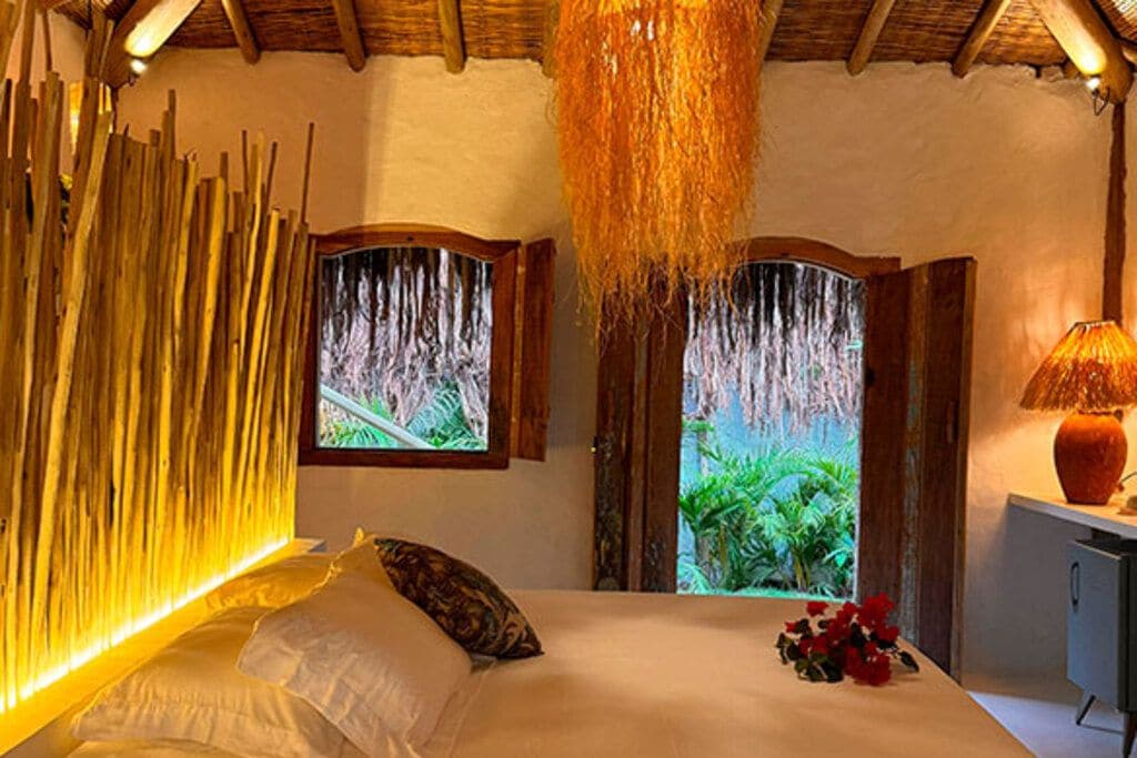 Quarto com cama, janela com vista para vegetação, planta pendurada e luminária.