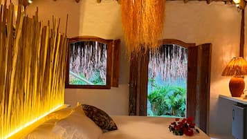 Quarto com cama, janela com vista para vegetação, planta pendurada e luminária.