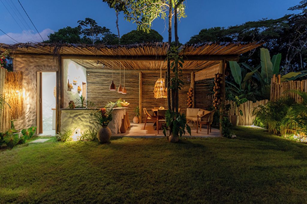 Área externa bem iluminada com mesa e cadeiras para refeições, cercada por plantas e um muro de madeira.