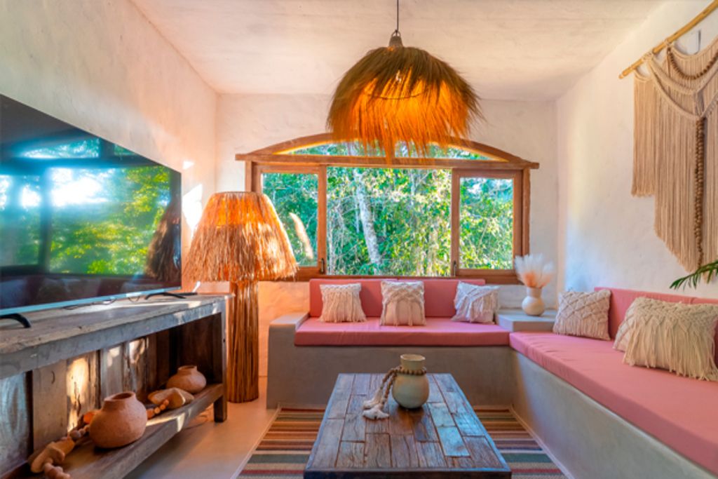 Um interior acolhedor com uma mesa de madeira, um sofá com almofadas, uma luminária pendente e uma janela com vista para o verde.