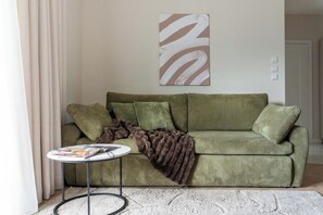 Um sofá verde com manta marrom, uma mesinha redonda com revistas e um quadro abstrato grande na parede.