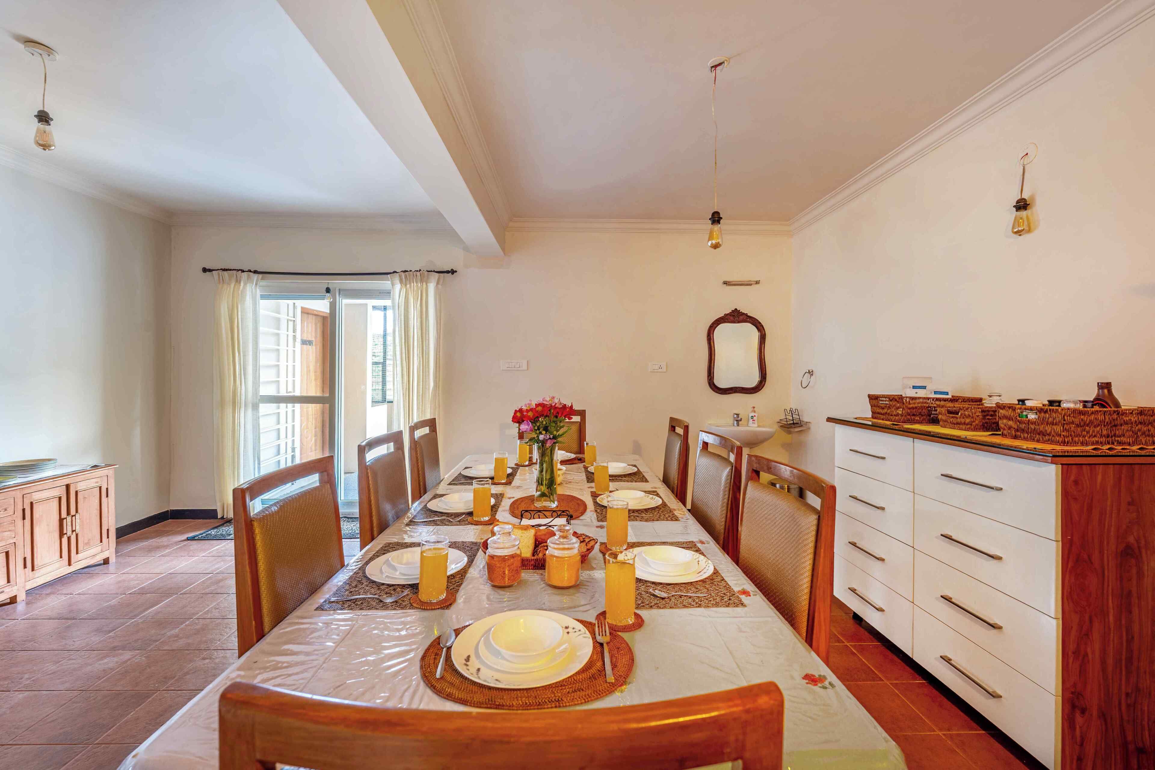 Uma sala de jantar com uma mesa comprida posta para refeição, cadeiras, um armário com gavetas e um espelho na parede.
