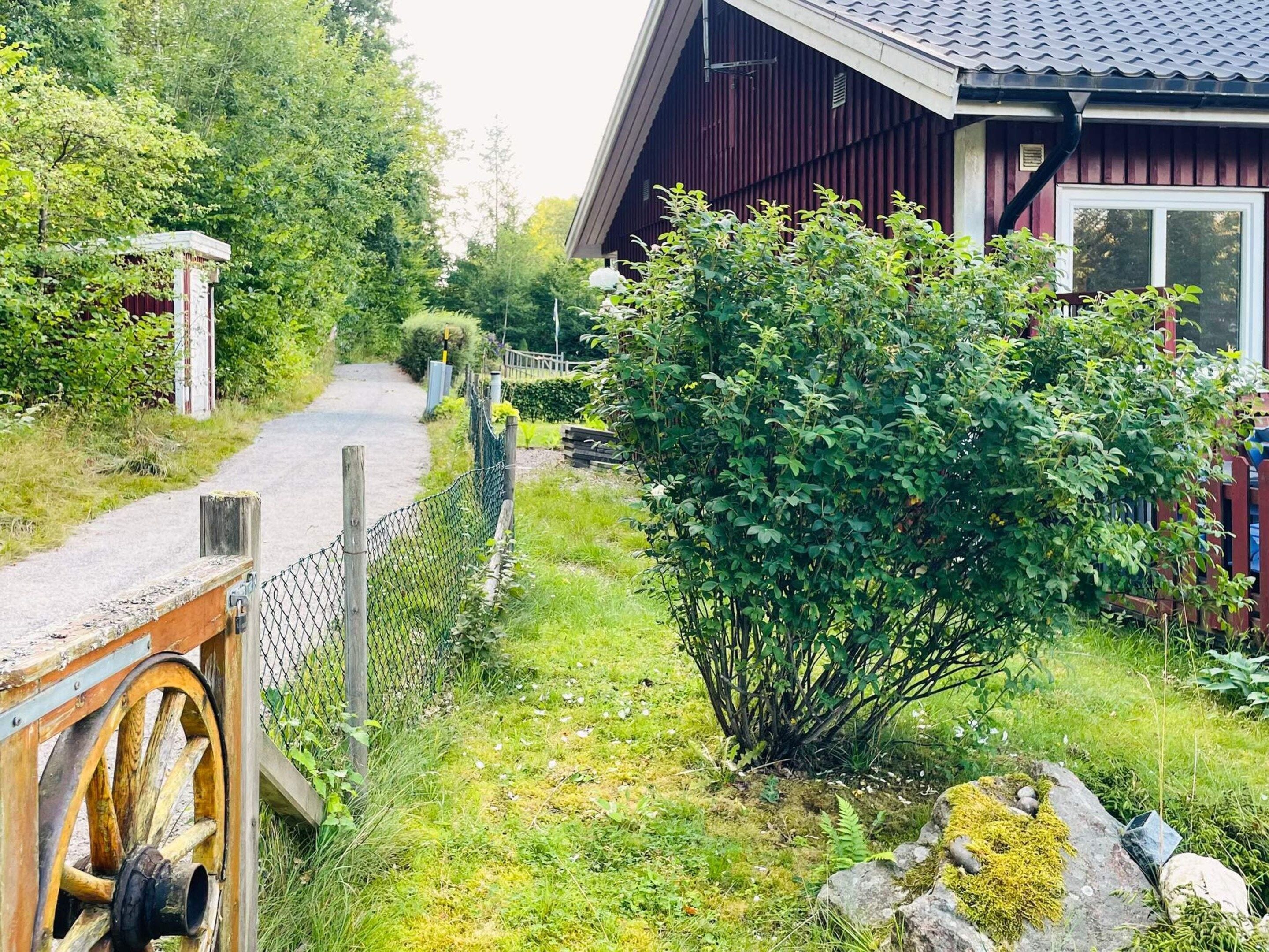 Uma propriedade rural com um acesso pavimentado que leva a um portão de madeira, cercada por vegetação exuberante e um edifício vermelho com telhado escuro.