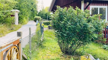 Uma propriedade rural com um acesso pavimentado que leva a um portão de madeira, cercada por vegetação exuberante e um edifício vermelho com telhado escuro.