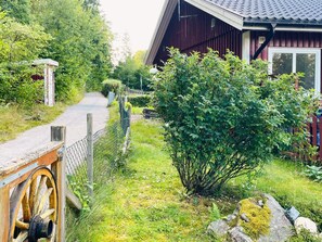 Uma propriedade rural com um acesso pavimentado que leva a um portão de madeira, cercada por vegetação exuberante e um edifício vermelho com telhado escuro.
