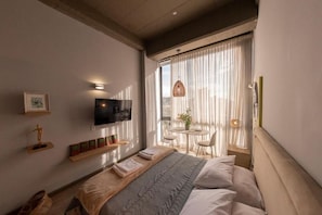 Quarto de hotel com cama, televisão na parede, mesa pequena com cadeiras e janela com cortinas.
