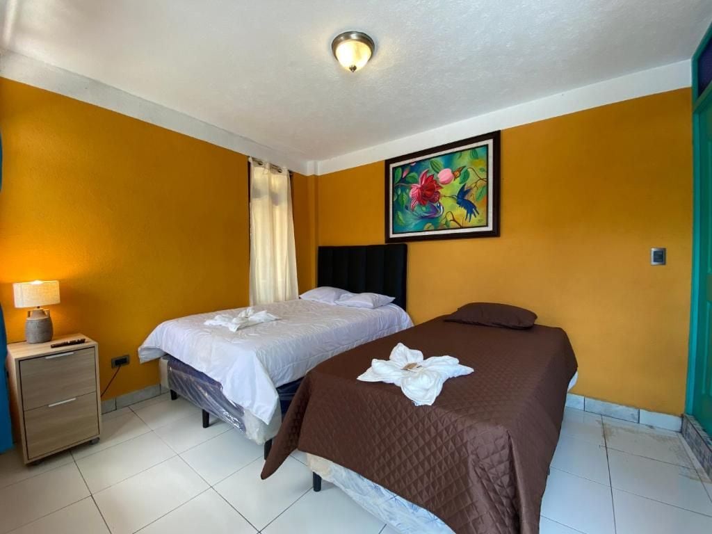 Quarto de hotel com duas camas, uma mesinha de cabeceira, um quadro na parede e uma janela com cortinas.