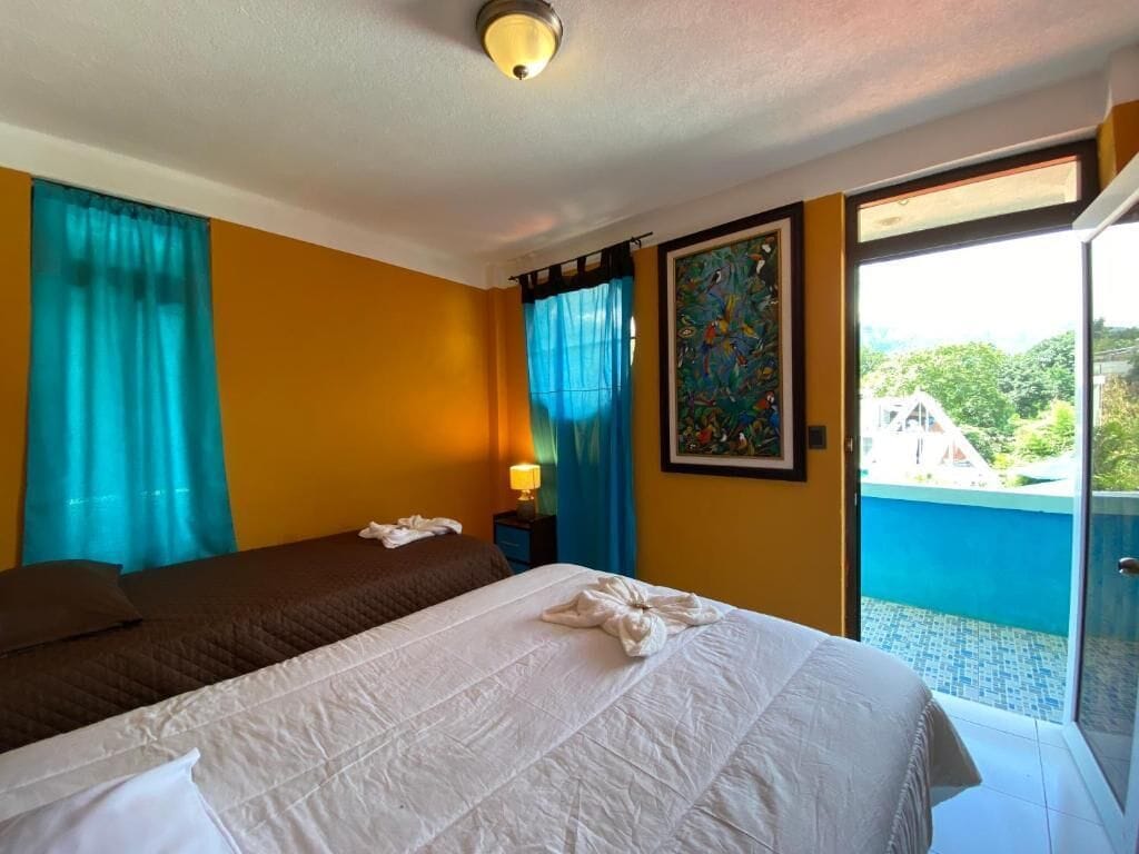 Quarto de hotel com cama, mesa de cabeceira, janela com cortinas e vista para o verde lá fora.