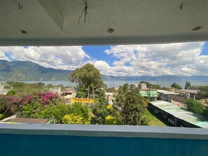 Vista de uma varanda com vista para um lago, montanhas ao longe, edifícios e árvores.