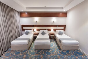 Quarto de hotel com três camas de solteiro, cabeceira de madeira, carpete estampado e uma grande janela com persianas.