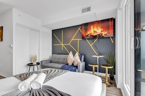 Quarto de hotel moderno com cama, sofá cinzento, mesinha e televisão na parede exibindo uma lareira.