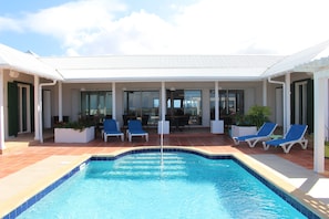 Área de piscina com piscina azul translúcida, rodeada por espreguiçadeiras e plantas em vasos.