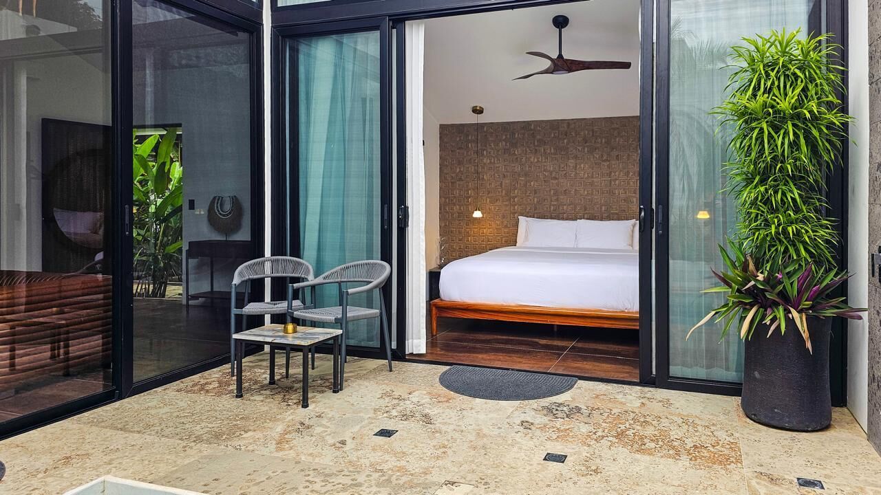 Quarto de hotel moderno com uma cama, área de estar com cadeiras, planta em vaso e porta de vidro que dá para uma área externa.