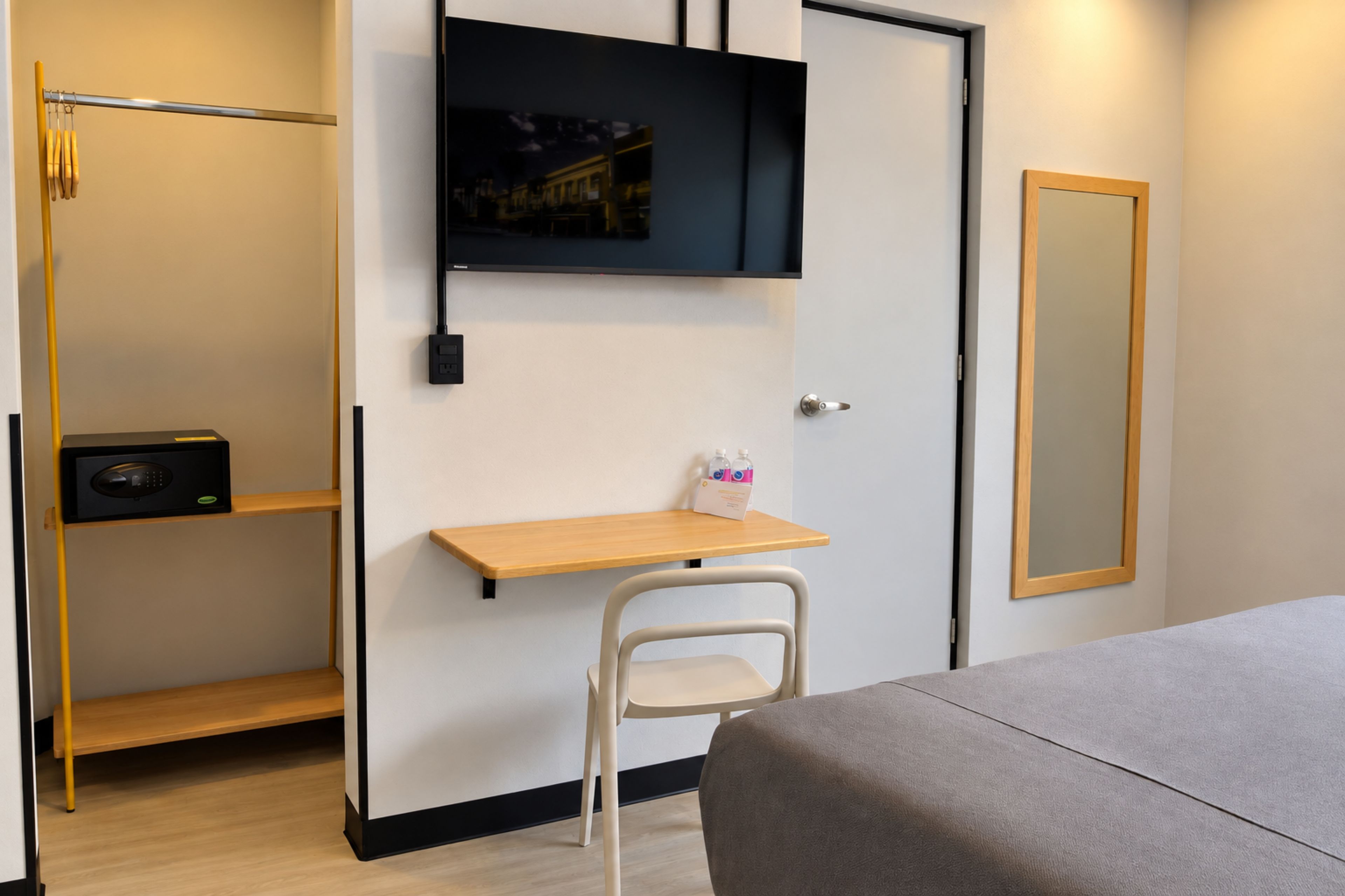 Quarto de hotel com cama, escrivaninha, cadeira, TV na parede, chuveiro e espelho.