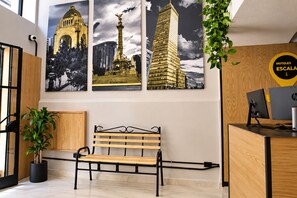 Uma área de recepção de hotel com um banco de madeira, uma mesa com computador e três fotografias emolduradas de paisagens urbanas na parede.
