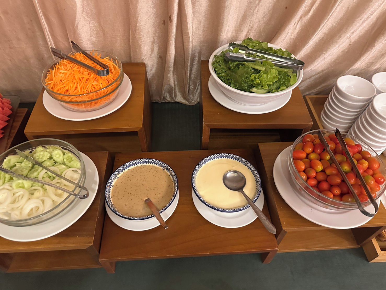 Mesa de buffet com diversos alimentos, incluindo cenoura ralada, alface, pepinos, sopa cremosa e tomate cereja.