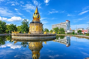 Uma fonte com estrutura central e estátuas ao redor, cercada por uma superfície aquática refletora, com um edifício ao fundo.