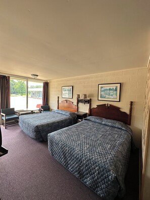 Quarto de hotel com duas camas, uma cadeira, uma mesa e uma janela com cortinas.