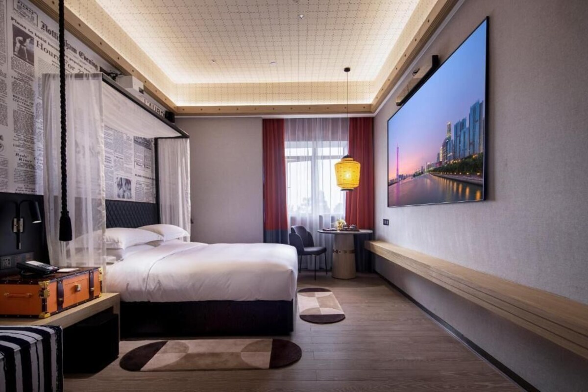 Quarto de hotel com uma cama grande, uma escrivaninha com luminária, uma cadeira, uma televisão e uma pintura com vista para a cidade.