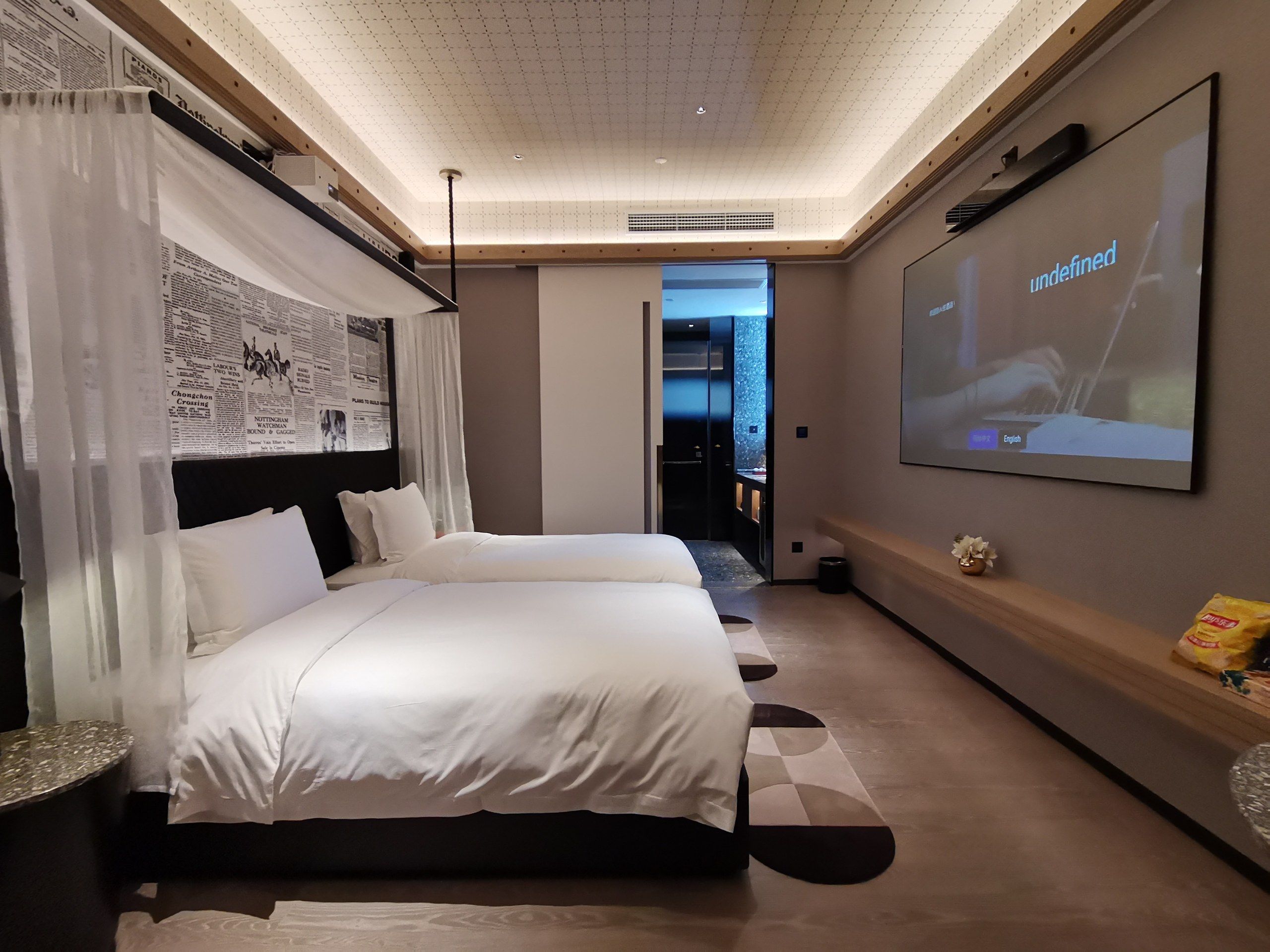 Quarto de hotel com duas camas, uma tela grande exibindo 'undefined' e uma parede com recortes de jornais.
