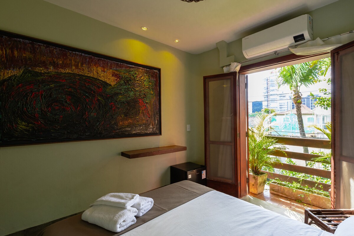 Quarto de hotel com cama, uma grande pintura abstrata e varanda com vista para prédios e áreas verdes.