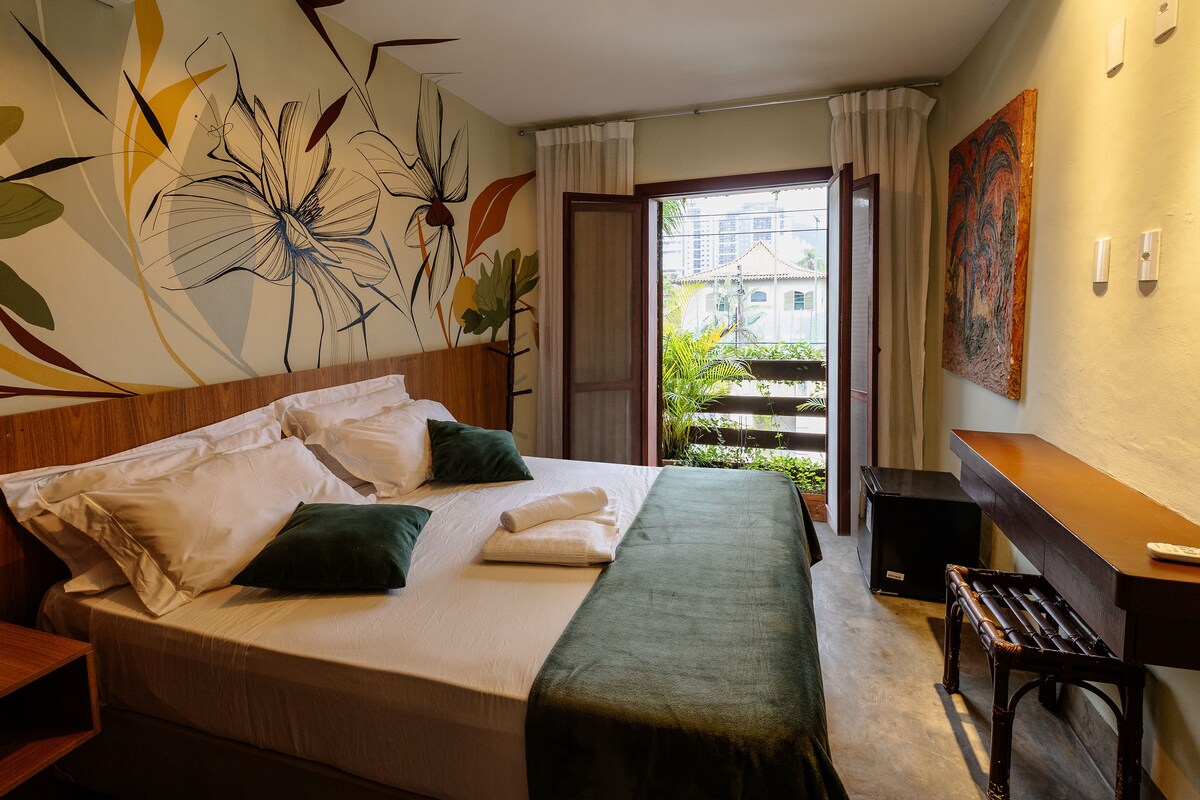 Quarto de hotel com cama, escrivaninha, cadeira, pintura e vista para prédios pela janela.