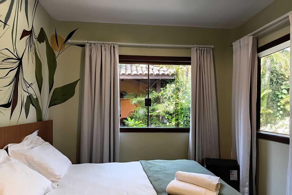 Quarto de hotel com cama, janela com cortinas e vista para o verde lá fora, além de uma parede com um mural de plantas.