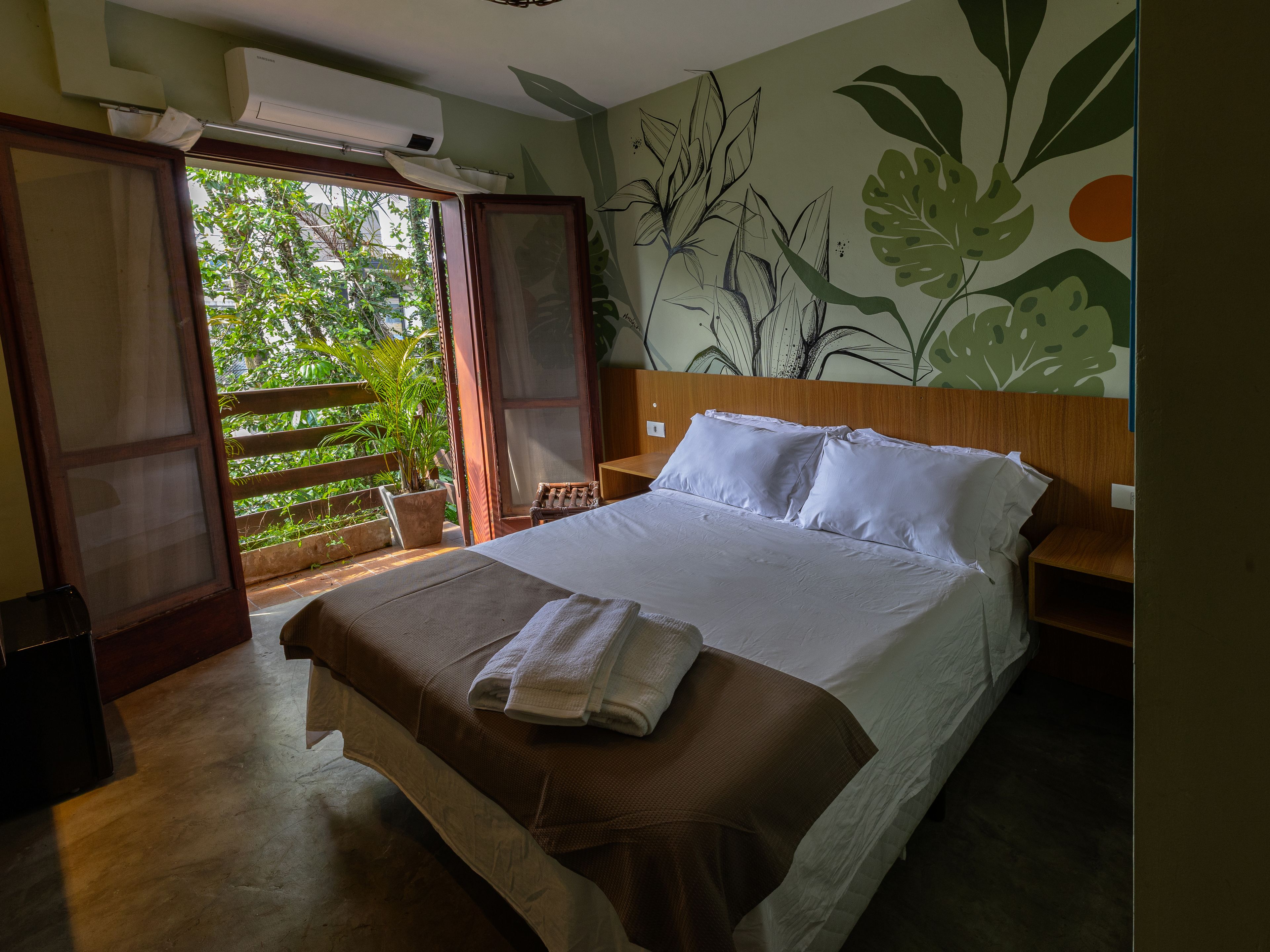 Quarto com cama, cabeceira de madeira, mesa de cabeceira e janela com vista para o verde.