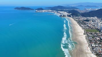 Vista aérea de uma cidade costeira com uma extensa praia, área urbana densa e montanhas ao longe.