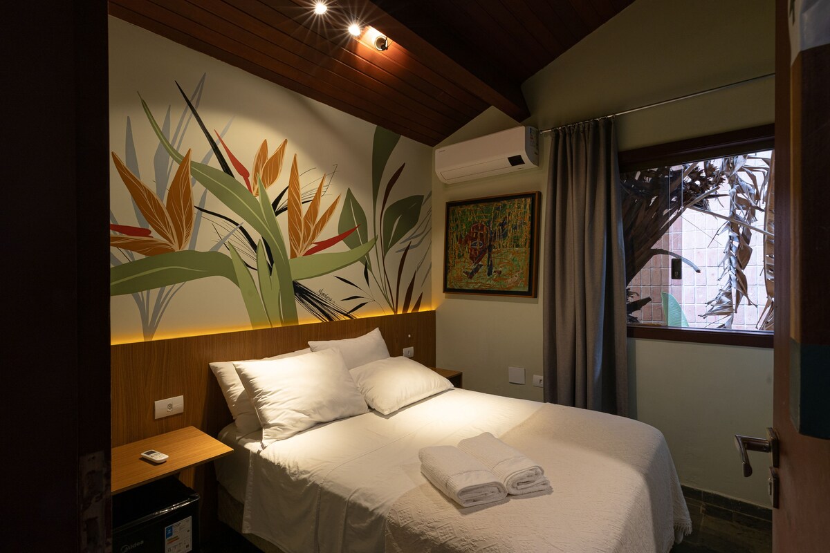 Quarto de hotel com cama, mesa de cabeceira, ar condicionado fixado na parede, um quadro e uma janela com cortinas.